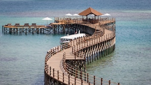 Um cais de madeira com um gazebo, mesas e guarda-sóis que se estende por um mar azul e límpido.