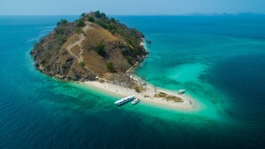 Vista aérea de uma pequena ilha com águas turquesa cristalinas, uma praia de areia e algumas embarcações.