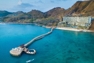 Vista aérea de um resort costeiro com um longo píer que se estende para o mar, um amplo edifício de hotel e montanhas ao fundo.