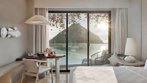 Quarto de hotel com cama, escrivaninha, cadeira, mesa com frutas e vista para montanha e água.