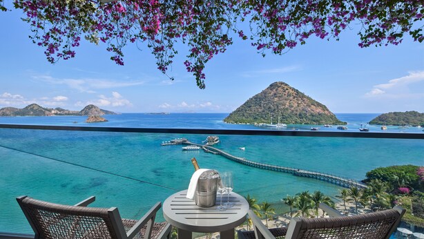Varanda com cadeiras de vime e uma mesa, com vista para o mar turquesa, barcos e um cais.