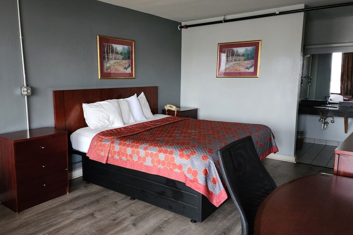 Quarto de hotel com cama de madeira, mesa de cabeceira, escrivaninha e banheiro com pia e espelho.