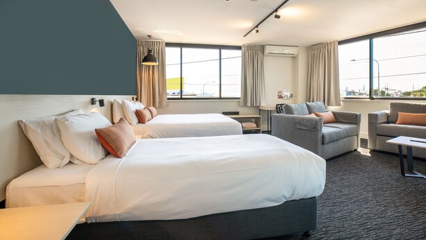 Quarto de hotel com duas camas, um sofá, uma mesa pequena e uma janela grande com vista para a cidade.