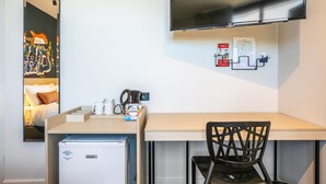 Quarto de hotel pequeno, com cama, escrivaninha e cadeira, mini-geladeira, televisão e cafeteira.
