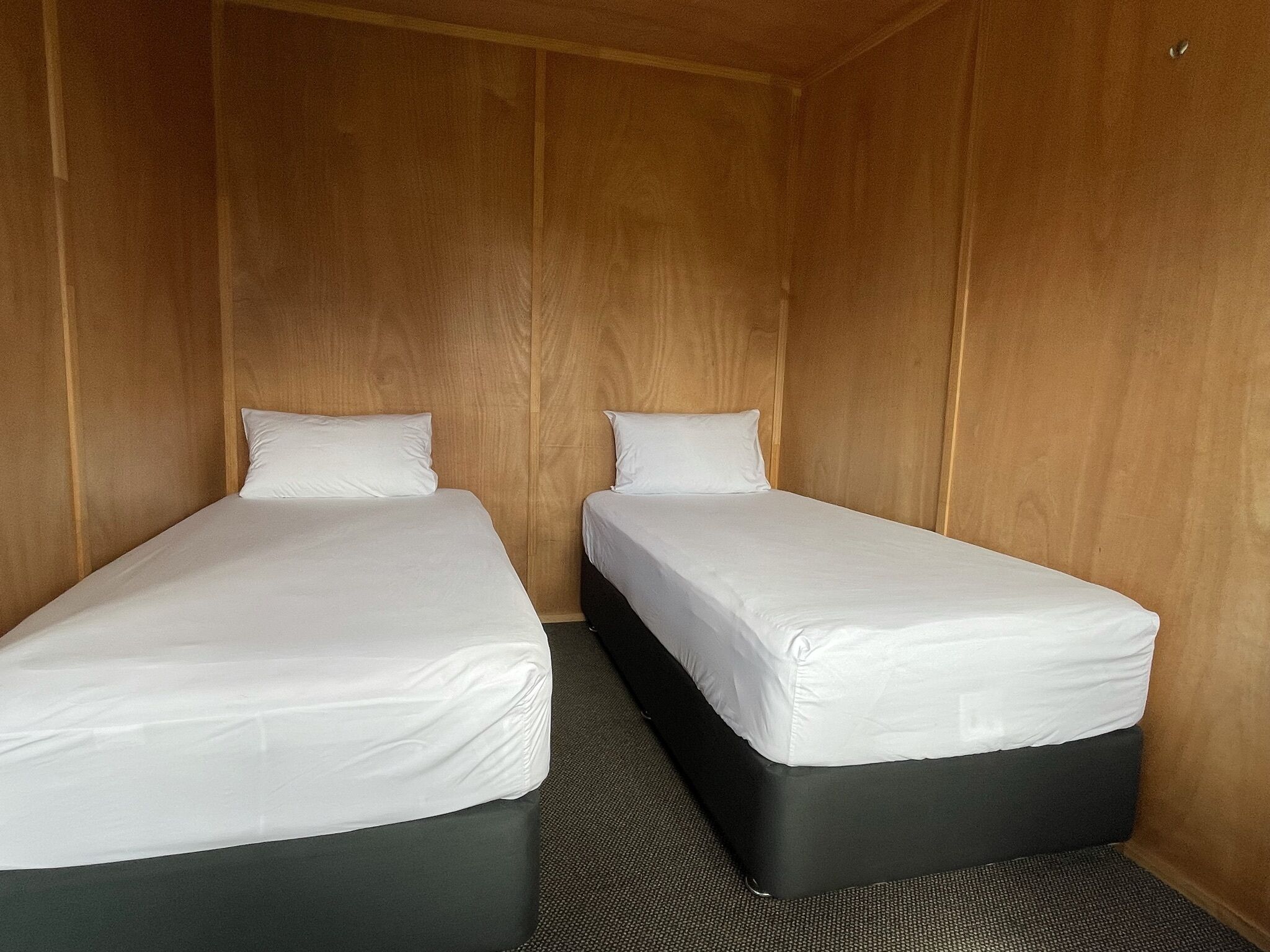 Um quarto pequeno de madeira com duas camas simples, cada uma com base preta e roupa de cama branca.