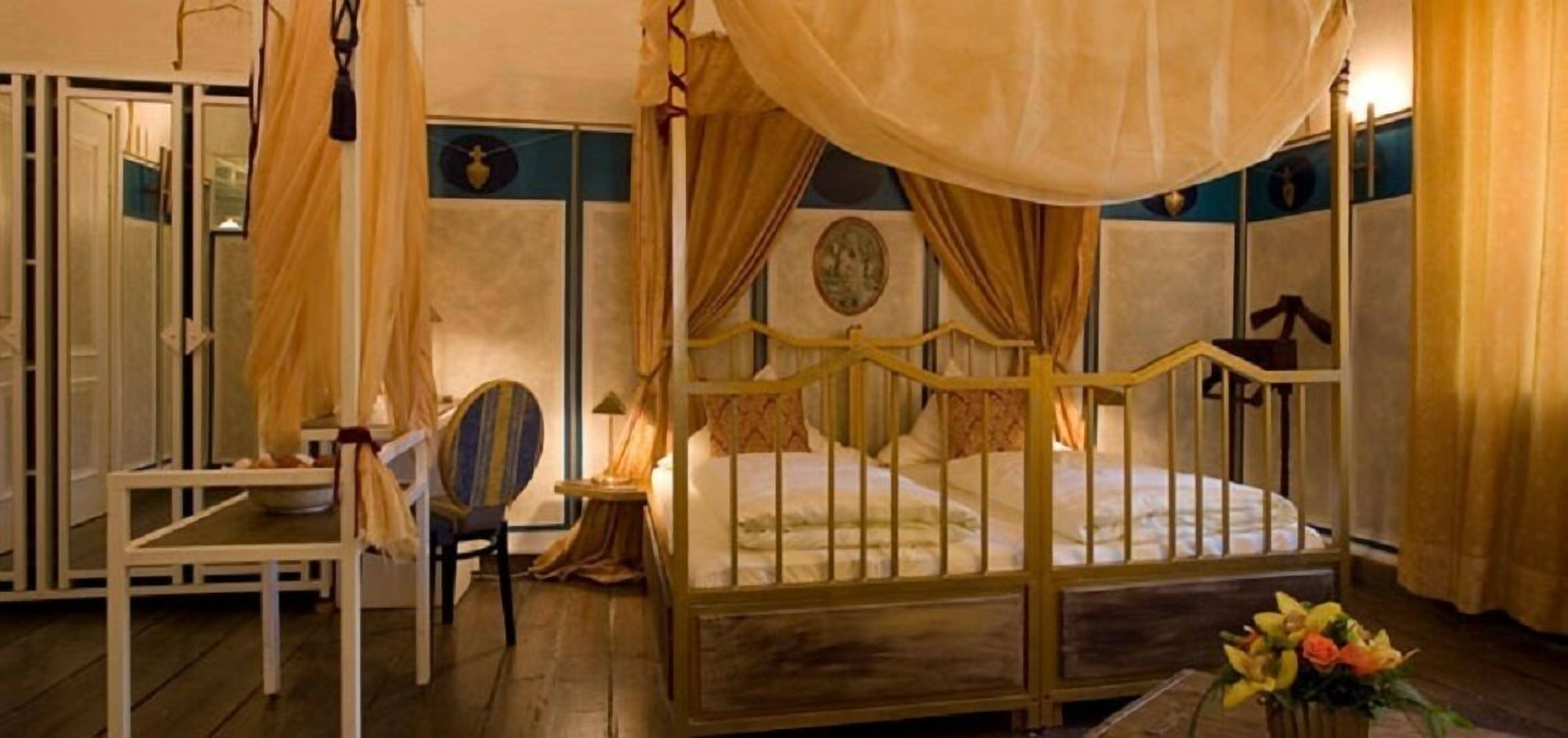 Uma cama com dossel e cortinas penduradas, estrutura de madeira, uma mesa de cabeceira com abajur e uma cadeira com almofada listrada.