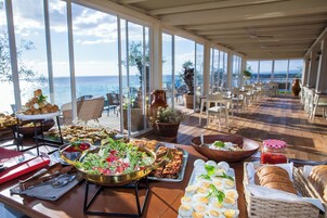 Mesa de buffet com diversos pratos, vista para o mar através de amplas janelas e um espaço de refeições informal com cadeiras e mesas.