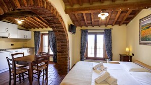Quarto de hotel com teto de madeira, cama com roupa de cama branca, mesa de jantar com cadeiras e cozinha com armários e pia.