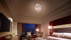 Quarto de hotel com uma cama grande, uma mesa com cadeira, televisão e vista para a cidade à noite.