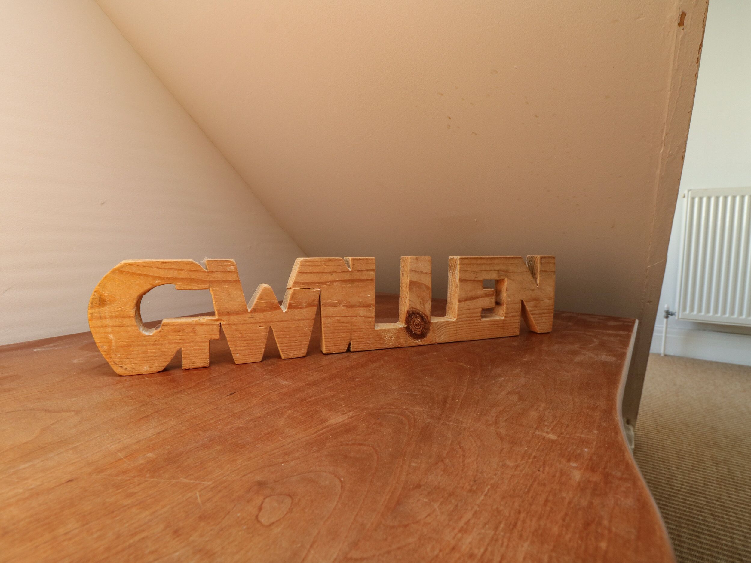 Letras de madeira formando a palavra 'GWILEN' sobre um piso de madeira.