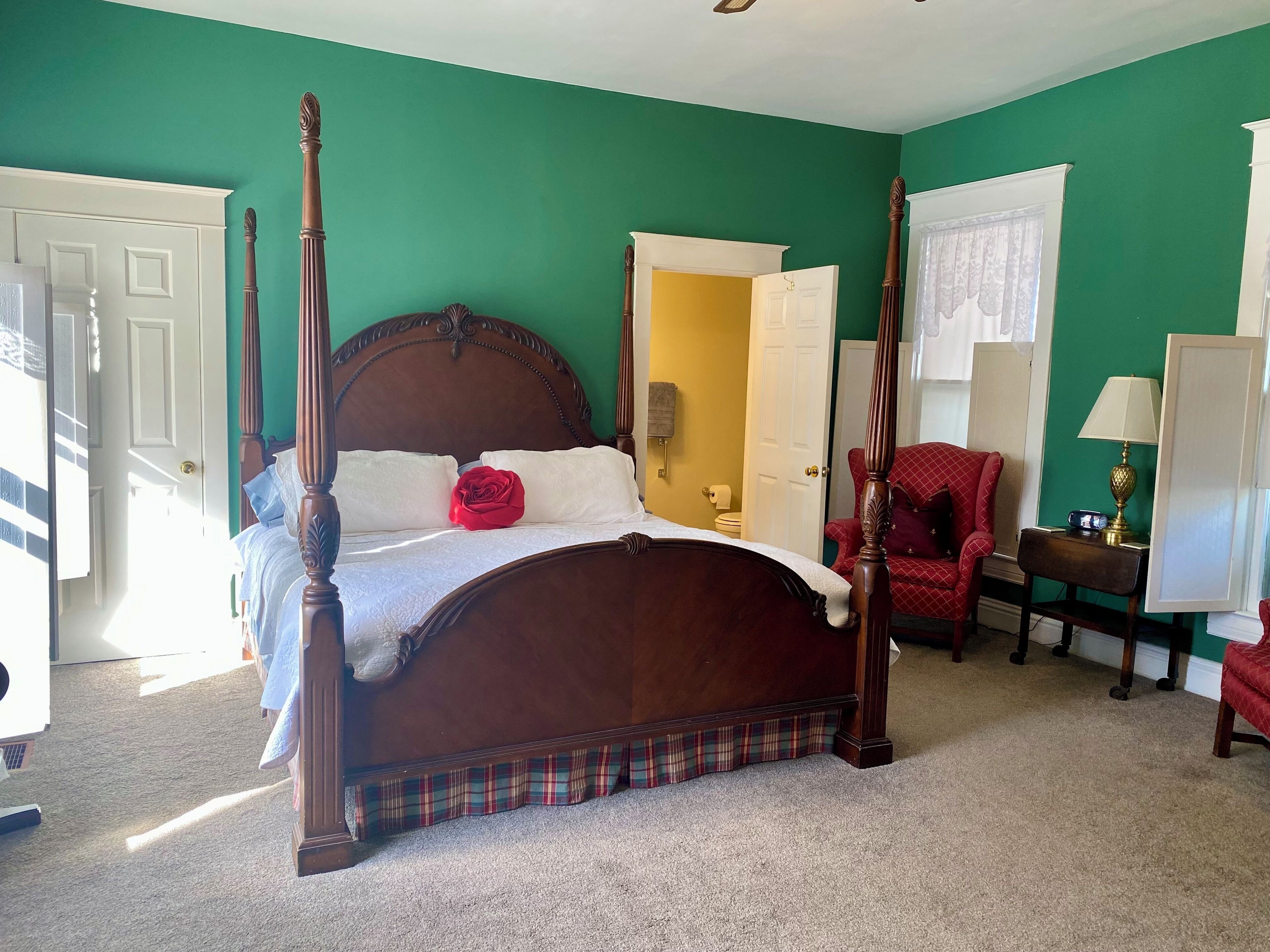 Uma cama com dossel, saia xadrez, travesseiro de rosa vermelha e edredom branco, num quarto com paredes verdes e porta branca.