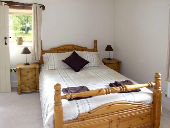 Uma cama de madeira com dossel, coberta por um edredom listrado em branco e cinza, com duas almofadas escuras e cabeceira de madeira. Há duas mesas de cabeceira com abajures, um criado-mudo de madeira e uma janela com cortinas.
