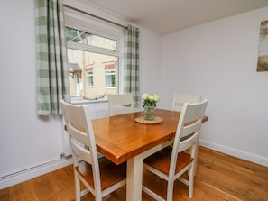 Uma sala de jantar com mesa de madeira e cadeiras brancas, um vaso com flores e uma janela com cortinas xadrez.