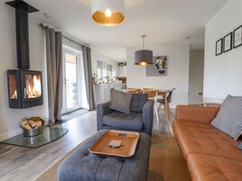Uma sala de estar moderna com uma lareira a lenha, sofás cinzentos, uma mesa de centro de madeira e uma área de jantar ao fundo.