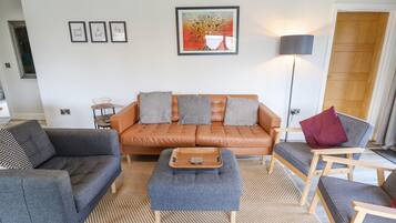 Sala de estar com um sofá de couro marrom, poltronas cinzas, um pequeno banquinho e um abajur. Há quadros nas paredes e uma porta de madeira ao fundo.