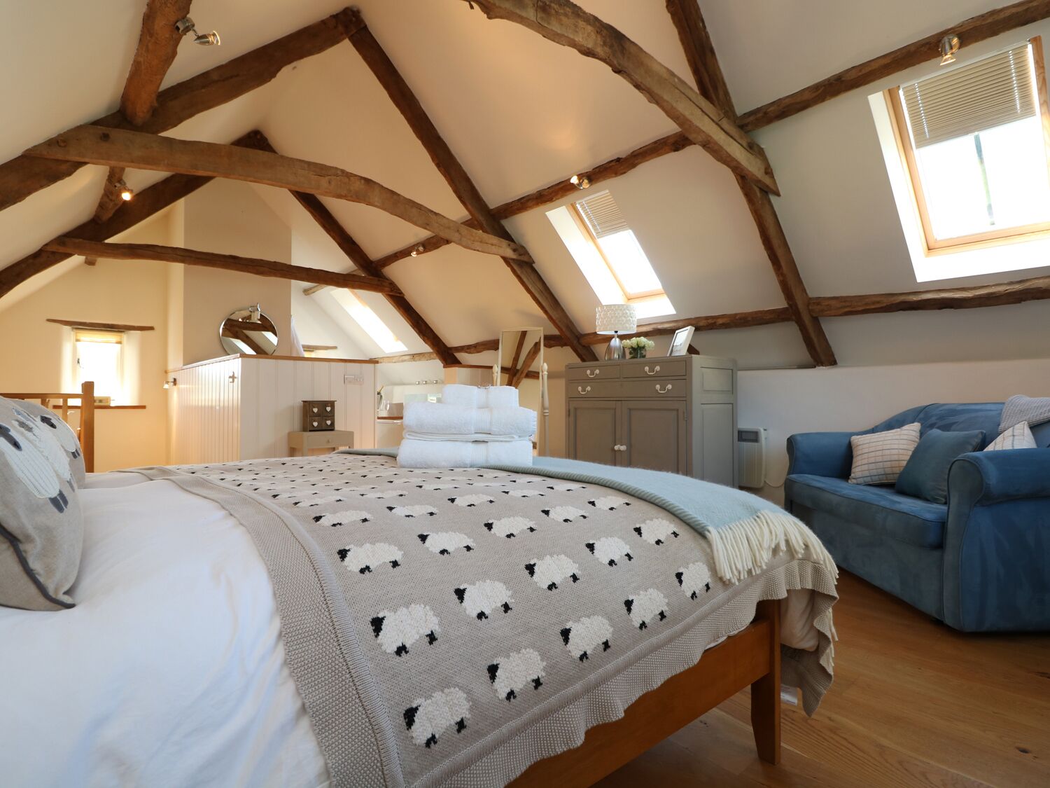 Quarto com teto inclinado, vigas de madeira, cama com manta estampada, sofá azul e cômoda.