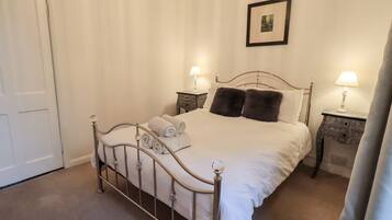 Cama bem arrumada com lençóis brancos, duas almofadas decorativas e uma pequena mesa de cabeceira com abajur.