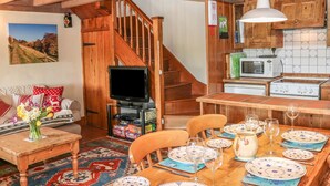 Uma sala de estar aconchegante com lareira, um espaço de jantar com mesa de madeira posta para quatro pessoas e uma cozinha com eletrodomésticos básicos.