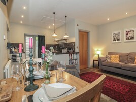 Uma área de refeições com uma mesa de madeira posta para dois, uma cozinha ao fundo e uma sala de estar com um sofá e um tapete vermelho estampado.