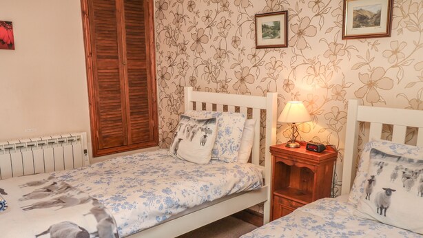 Quarto com duas camas, cada uma com roupa de cama florida e estampas de ovelhas, uma mesinha de cabeceira de madeira com abajur e um radiador.