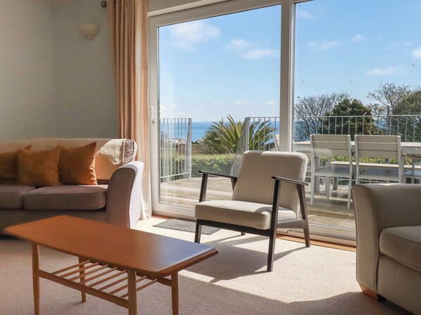 Sala de estar com sofá, mesa e cadeira, varanda com vista para o mar e mobiliário de exterior.
