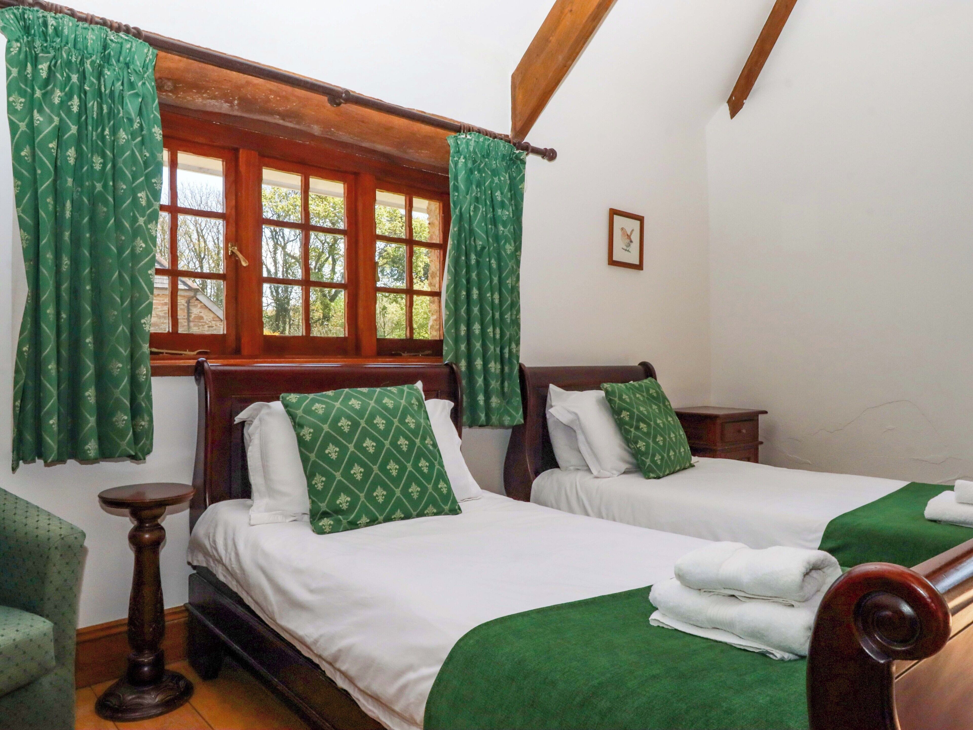 Quarto com duas camas, cabeceiras de madeira, janela com cortinas, uma mesinha e uma cadeira.