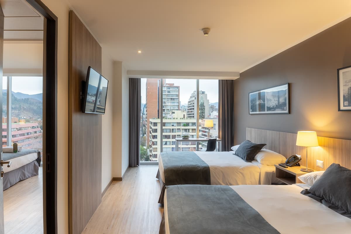Quarto de hotel com duas camas, televisão, escrivaninha com luminária e vista para a cidade.