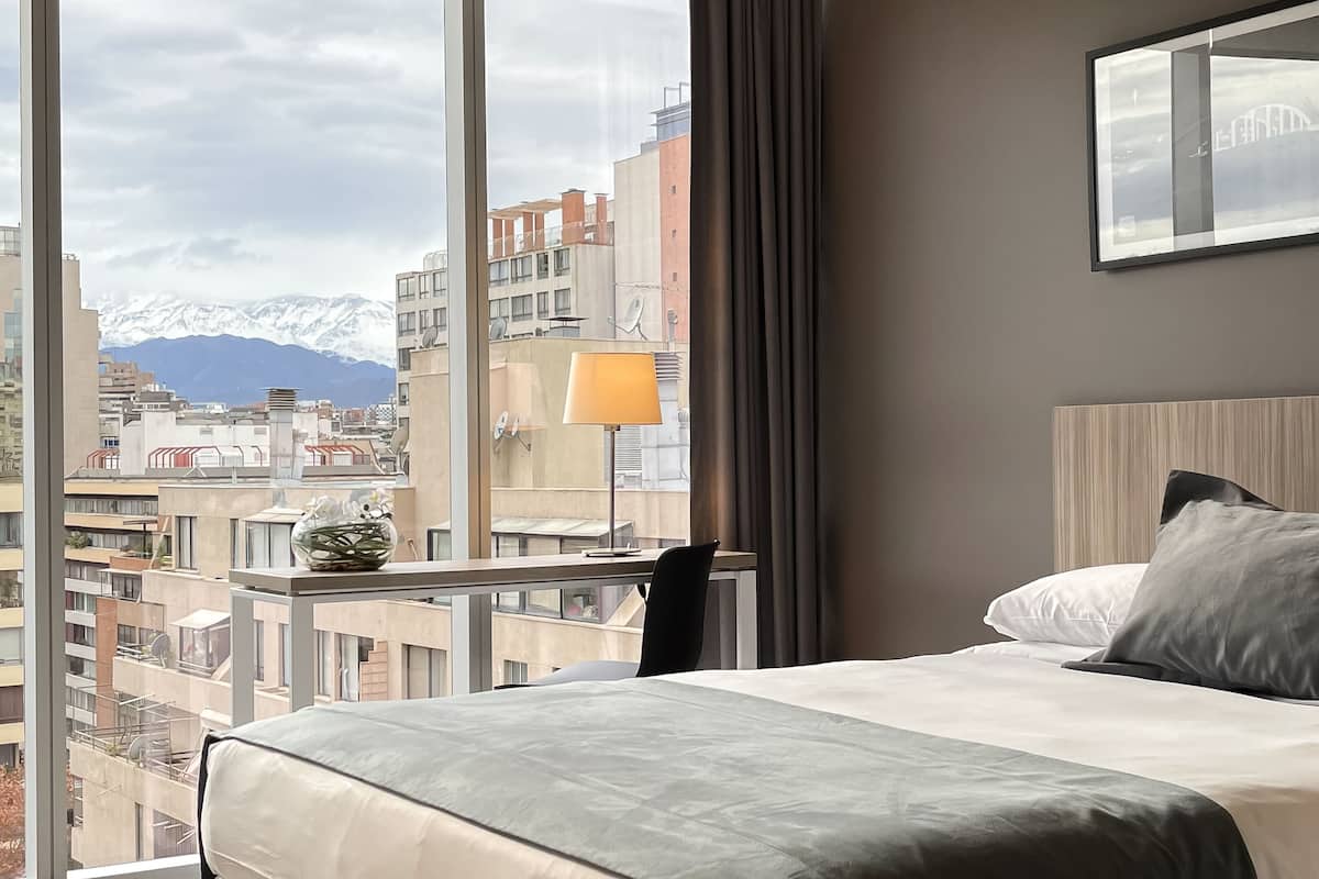 Quarto de hotel com uma grande janela que oferece vista para a cidade, uma cama com edredom cinza e branco, cabeceira de madeira e um quadro na parede.