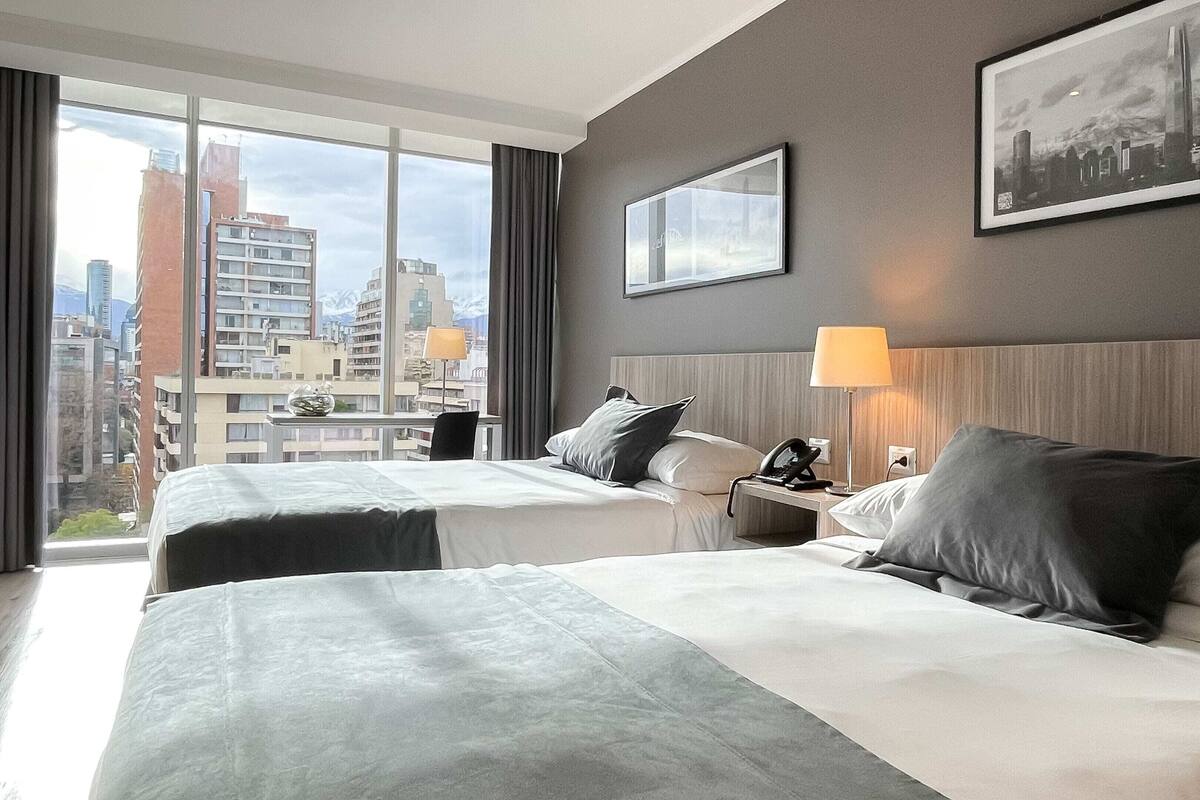 Quarto de hotel com duas camas, vista para a cidade, um quadro pendurado na parede e uma luminária na mesinha de cabeceira.