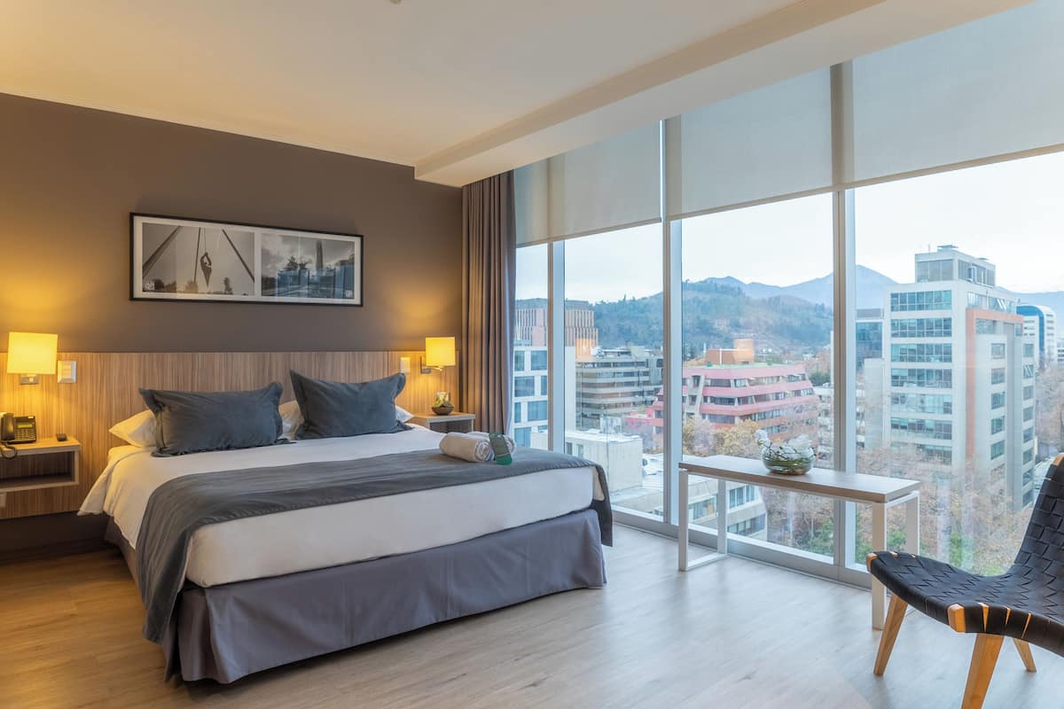 Quarto de hotel moderno com uma cama grande, duas luminárias de cabeceira, vista da cidade e uma pequena mesa com uma planta.