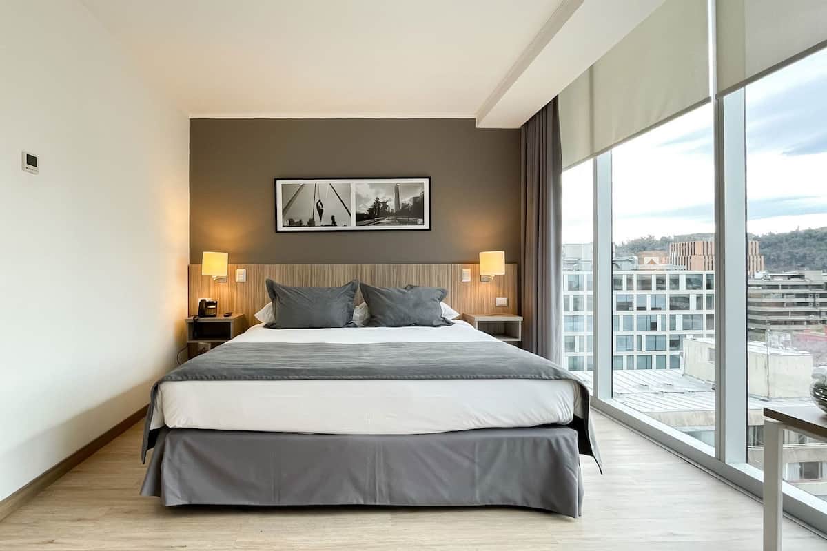 Quarto de hotel moderno com uma cama grande, abajures, vista para a cidade e um quadro na parede.