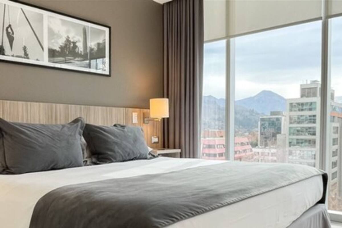 Quarto de hotel com uma cama grande, mesa de cabeceira, abajur e vista para a cidade através de amplas janelas.