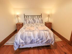 Uma cama bem arrumada com colcha estampada e duas toalhas dobradas, com mesas de cabeceira com abajures de cada lado, em um quarto com piso de madeira e tapete estampado.