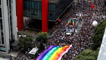 Uma grande multidão se reuniu em uma praça da cidade, com uma bandeira do arco-íris em destaque.