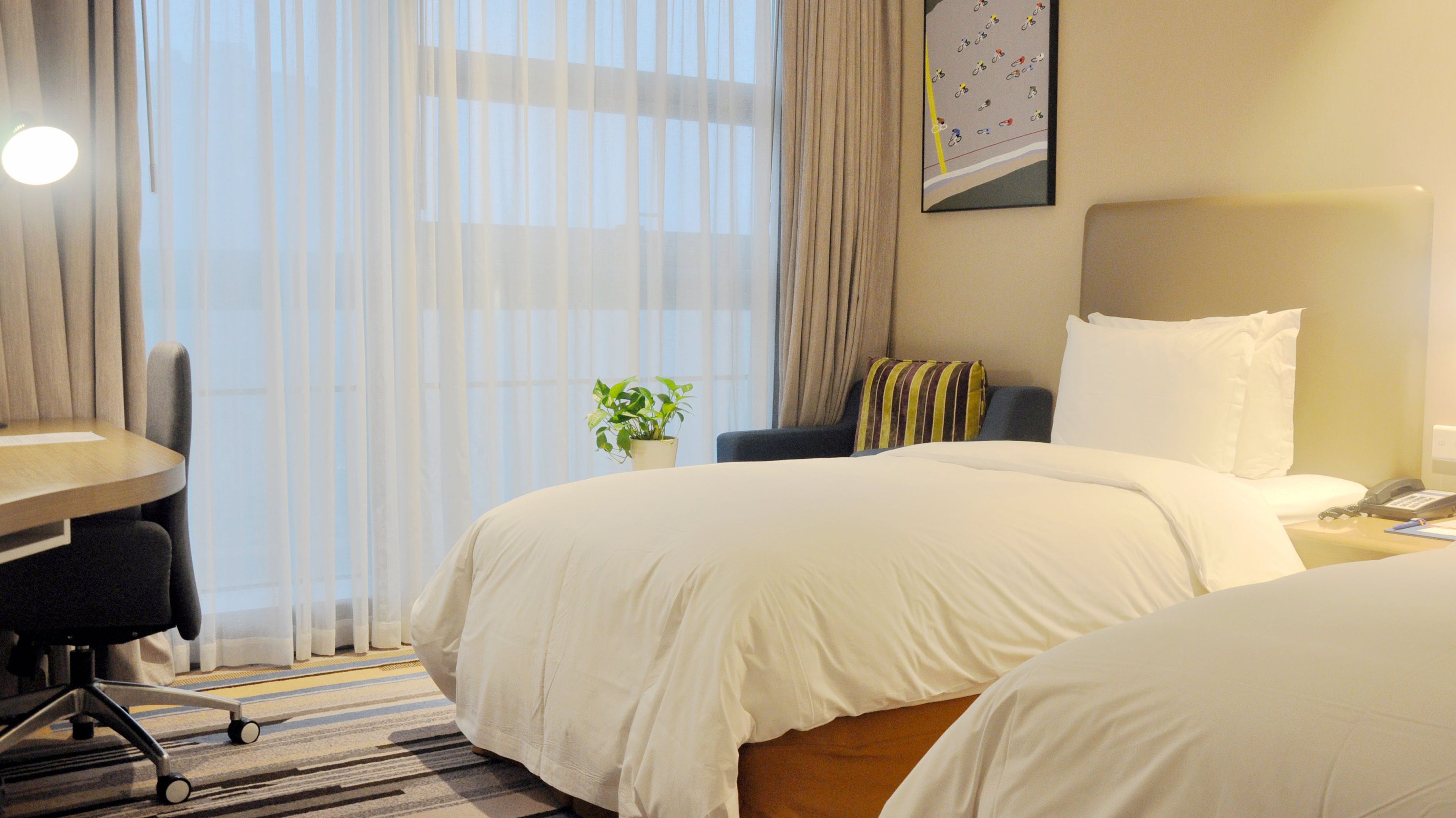 Quarto de hotel com duas camas, uma escrivaninha, uma cadeira, um vaso de planta e um quadro na parede.