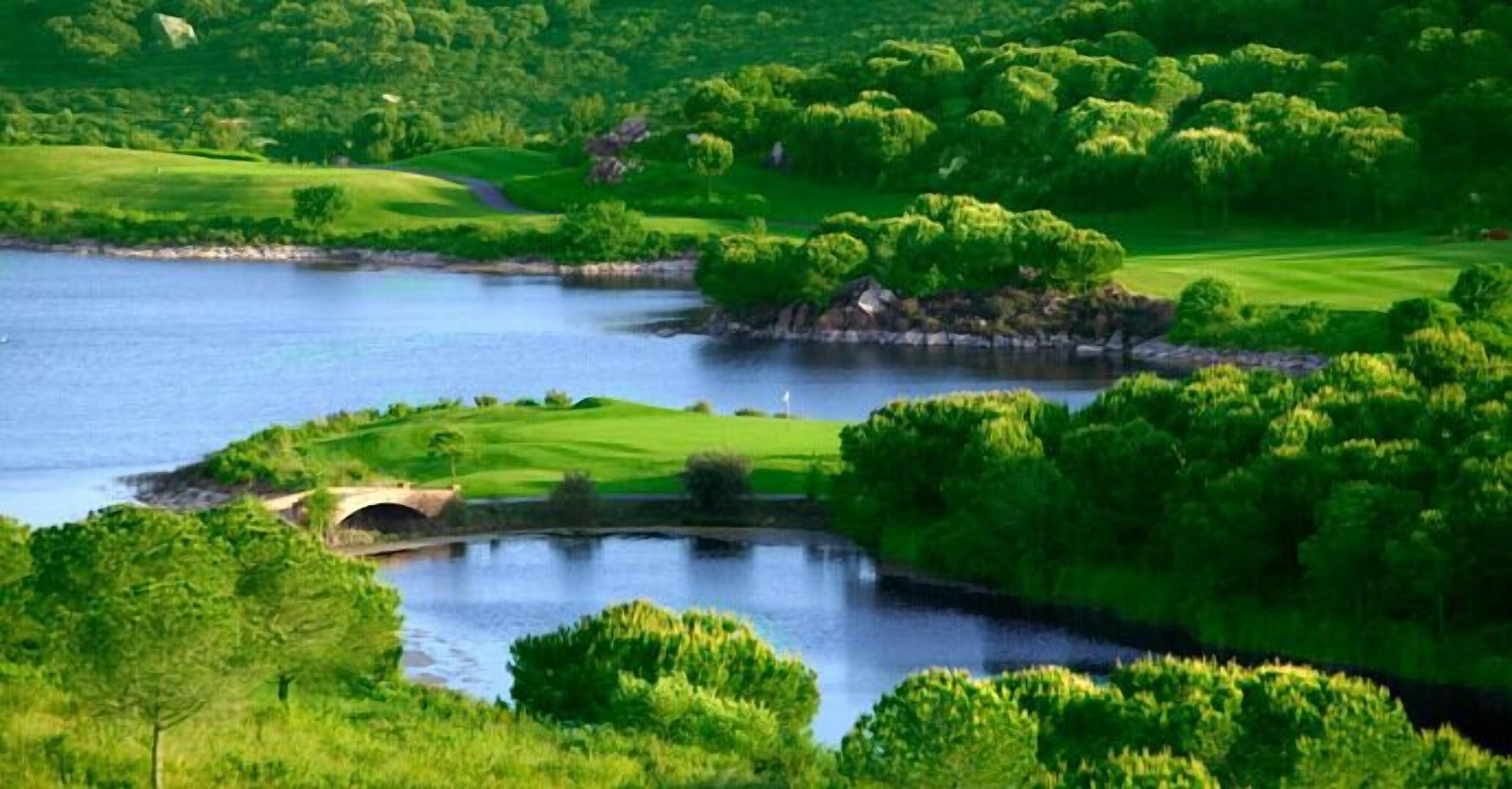 Um campo de golfe com um lago, vegetação exuberante e uma ponte.