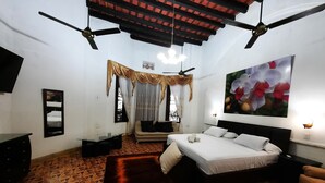 Quarto com cama, sofá, ventilador de teto, lustre e uma grande janela com cortinas.