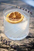 Um copo de vidro transparente com gelo e uma fatia de laranja seca, apoiado em uma superfície texturizada com raminhos de lavanda.
