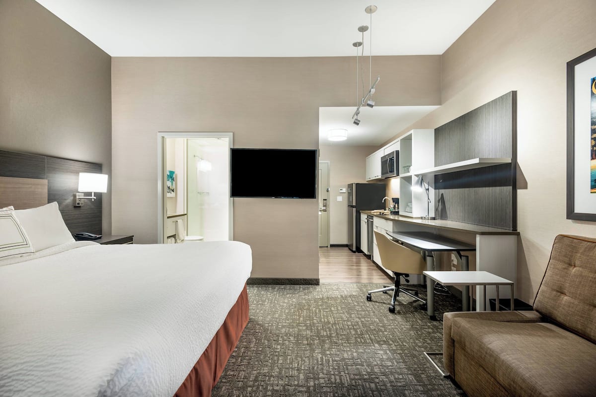 Quarto de hotel com uma cama grande, escrivaninha, sofá, televisão e cozinha compacta.