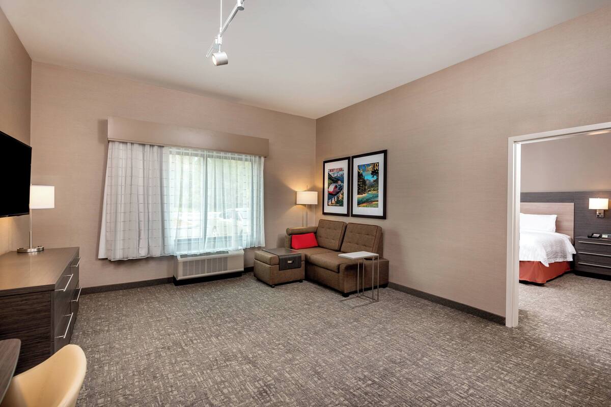 Quarto de hotel com sofá, escrivaninha, cadeira, cama, televisão e quadros nas paredes.