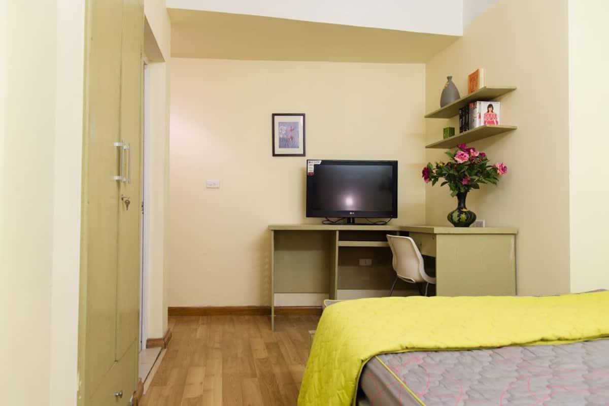 Quarto com cama, escrivaninha com televisão, cadeira e prateleira com livros e um vaso de flores.