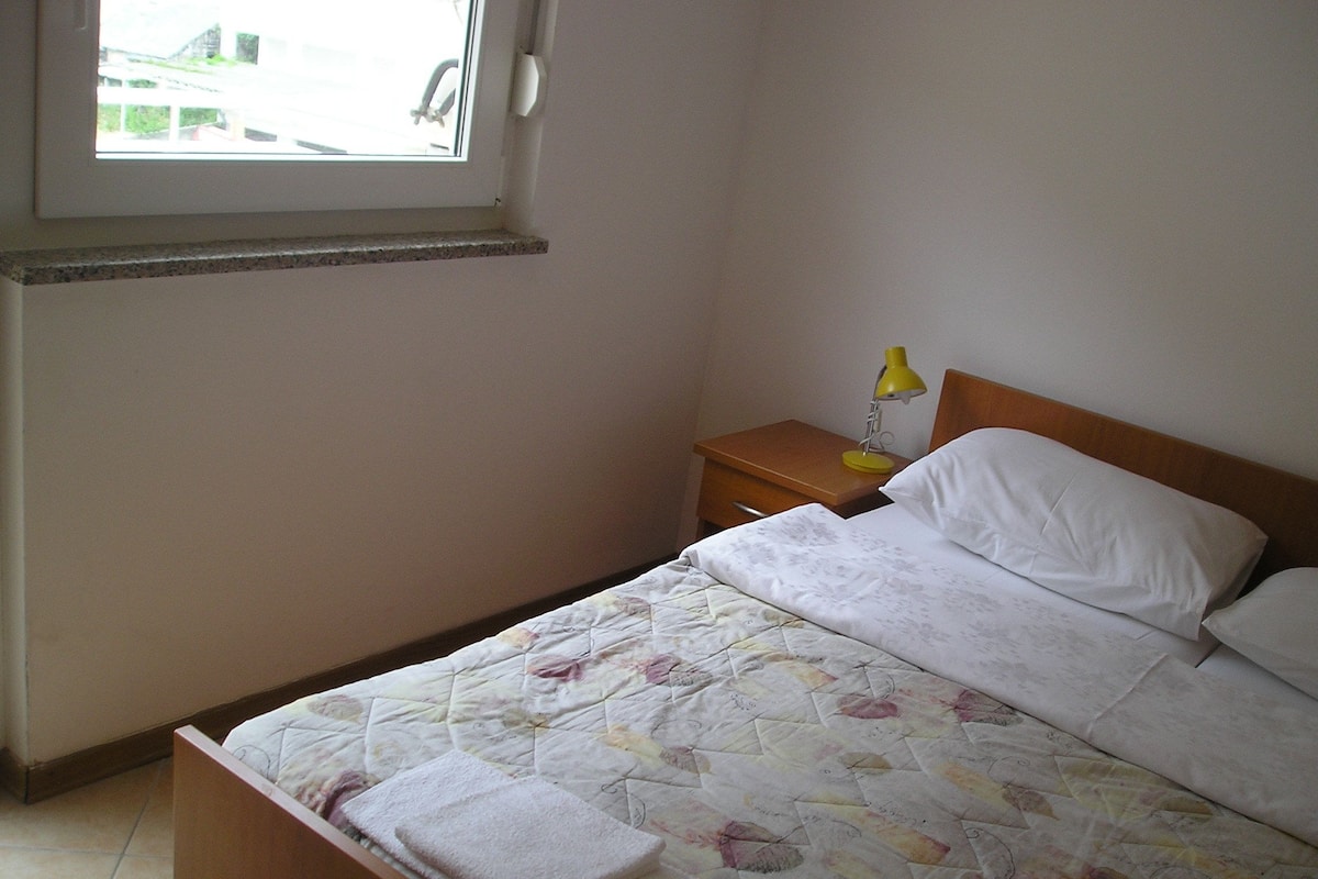 Uma cama bem arrumada com colcha florida, uma luminária de cabeceira e uma janela com vista para o exterior.