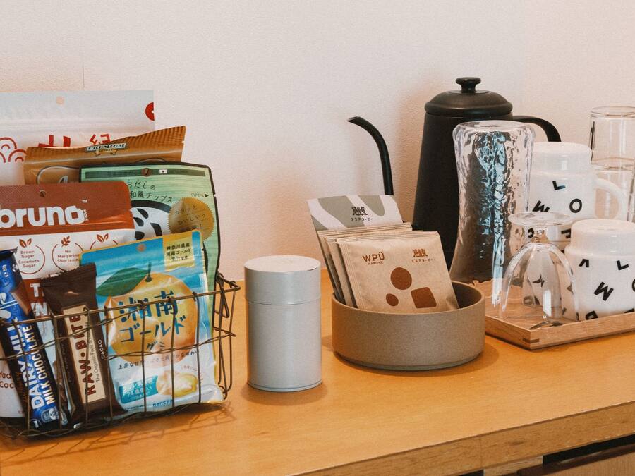 Uma bancada de cozinha com diversos utensílios, incluindo uma cafeteira, um bule, um moedor de café, um espumador de leite e uma bandeja com xícaras de café e um jarro de vidro.