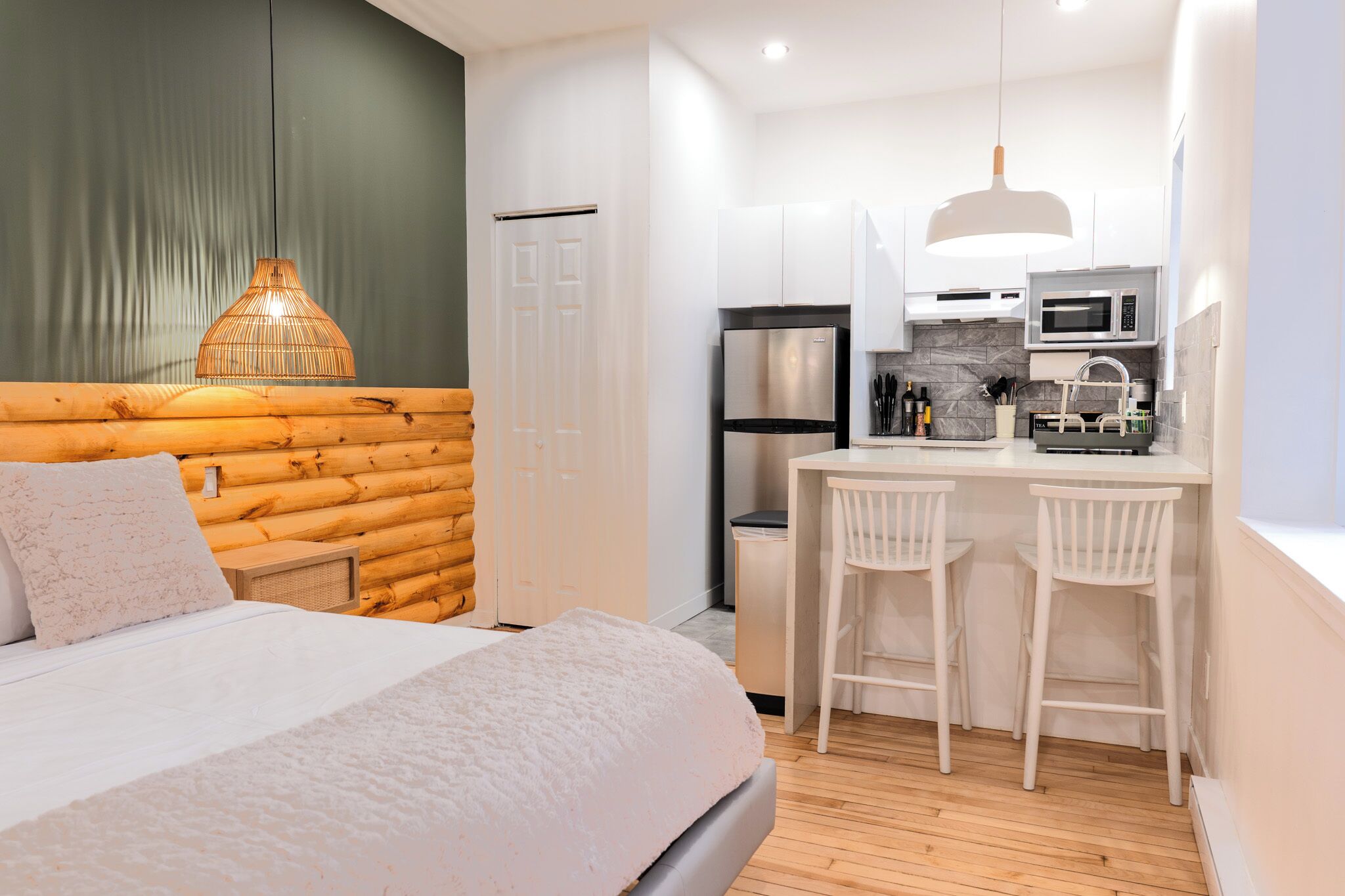 Quarto moderno com cabeceira de madeira, cama com edredom branco, mesa de cabeceira e luminária pendente. Anexo ao quarto, uma kitchenette compacta com geladeira, micro-ondas e banquetas.