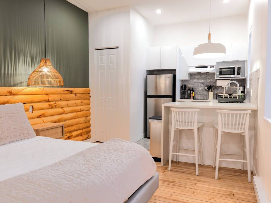 Quarto moderno com cabeceira de madeira, cama com edredom branco, mesa de cabeceira e luminária pendente. Anexo ao quarto, uma kitchenette compacta com geladeira, micro-ondas e banquetas.