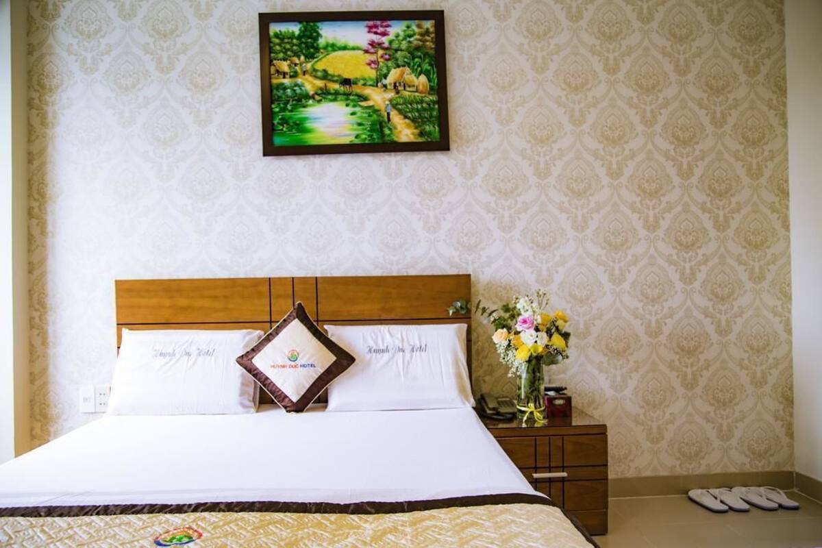 Cama bem arrumada com cabeceira de madeira, dois travesseiros e um travesseiro decorativo com um logotipo. Uma moldura com uma imagem está pendurada acima da cama, e um vaso com flores está em uma mesa de cabeceira ao lado da cama.