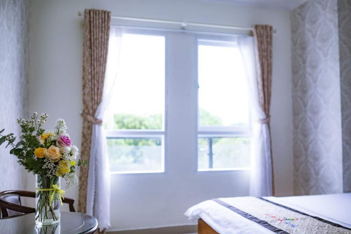 Quarto de hotel com uma cama, uma mesa com um vaso de flores e uma janela com cortinas.
