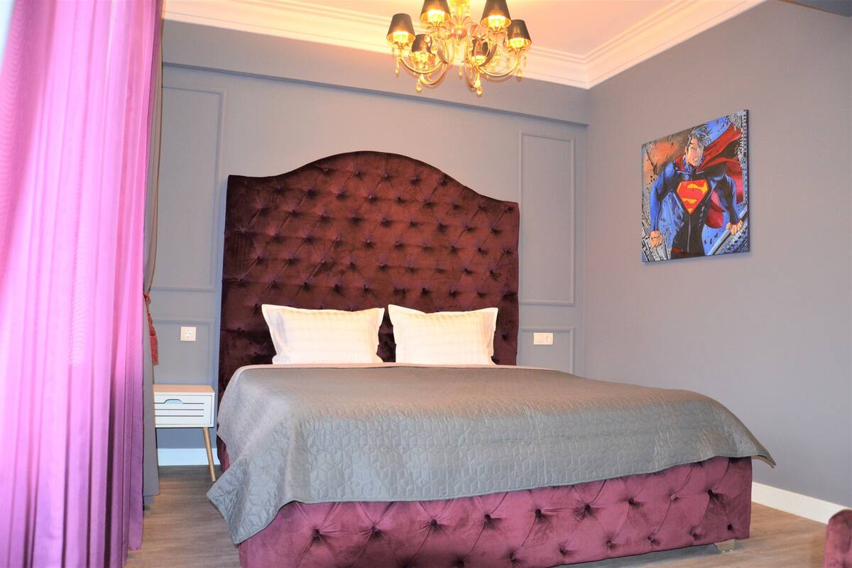 Quarto com cabeceira estofada, cama com colcha cinzenta e um quadro na parede.