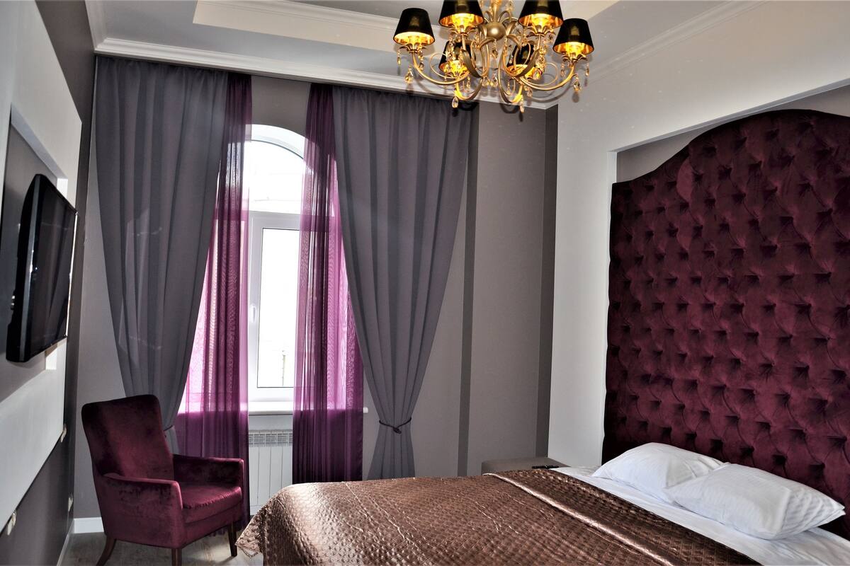 Quarto com uma cama grande, uma poltrona de veludo vermelha, um candelabro e uma televisão na parede.