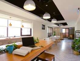 Um espaço de escritório moderno com escrivaninhas de madeira, laptops e livros. Janelas amplas permitem a entrada de luz natural, e o ambiente possui pé direito alto com iluminação pendente.